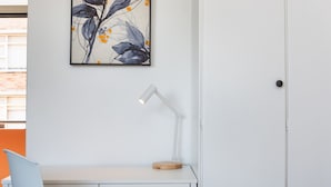 Quarto moderno com escrivaninha branca, luminária branca e quadro com flores na parede.
