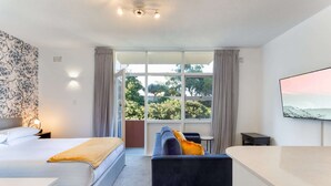 Quarto de hotel com cama, sofá, escrivaninha e uma janela grande com vista para o verde.