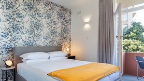 Quarto com cama, mesa de cabeceira, parede com papel de parede floral e vista para o verde lá fora.