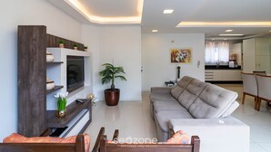 Uma sala de estar moderna com um sofá cinzento, uma TV sobre um suporte de madeira, uma planta em vaso e uma área de jantar ao fundo.