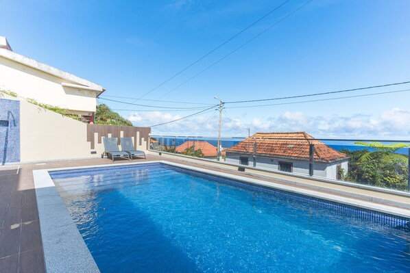 Piscina com água azul cristalina, cercada por um deck ladrilhado. Há espreguiçadeiras e um pequeno edifício com telhado telhado por perto.