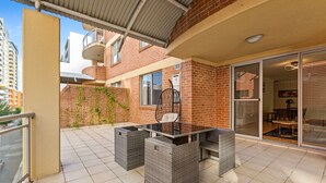 Uma varanda com móveis de vime, uma mesa de vidro e vista para um edifício de tijolos.