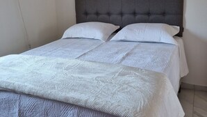 Uma cama de casal com cabeceira cinza revestida, roupa de cama branca e parede lisa.