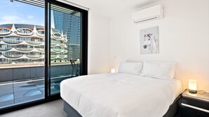 Quarto de hotel moderno com uma cama grande, mesa de cabeceira, ar condicionado fixado na parede e uma janela com vista para a cidade.