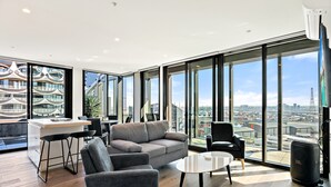 Uma sala de estar moderna com sofá, cadeiras, uma pequena mesa e uma escrivaninha com computador. Janelas amplas proporcionam vista para a cidade.