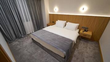 Quarto de hotel com uma cama, duas mesas de cabeceira, um criado-mudo, uma lâmpada e uma cortina.