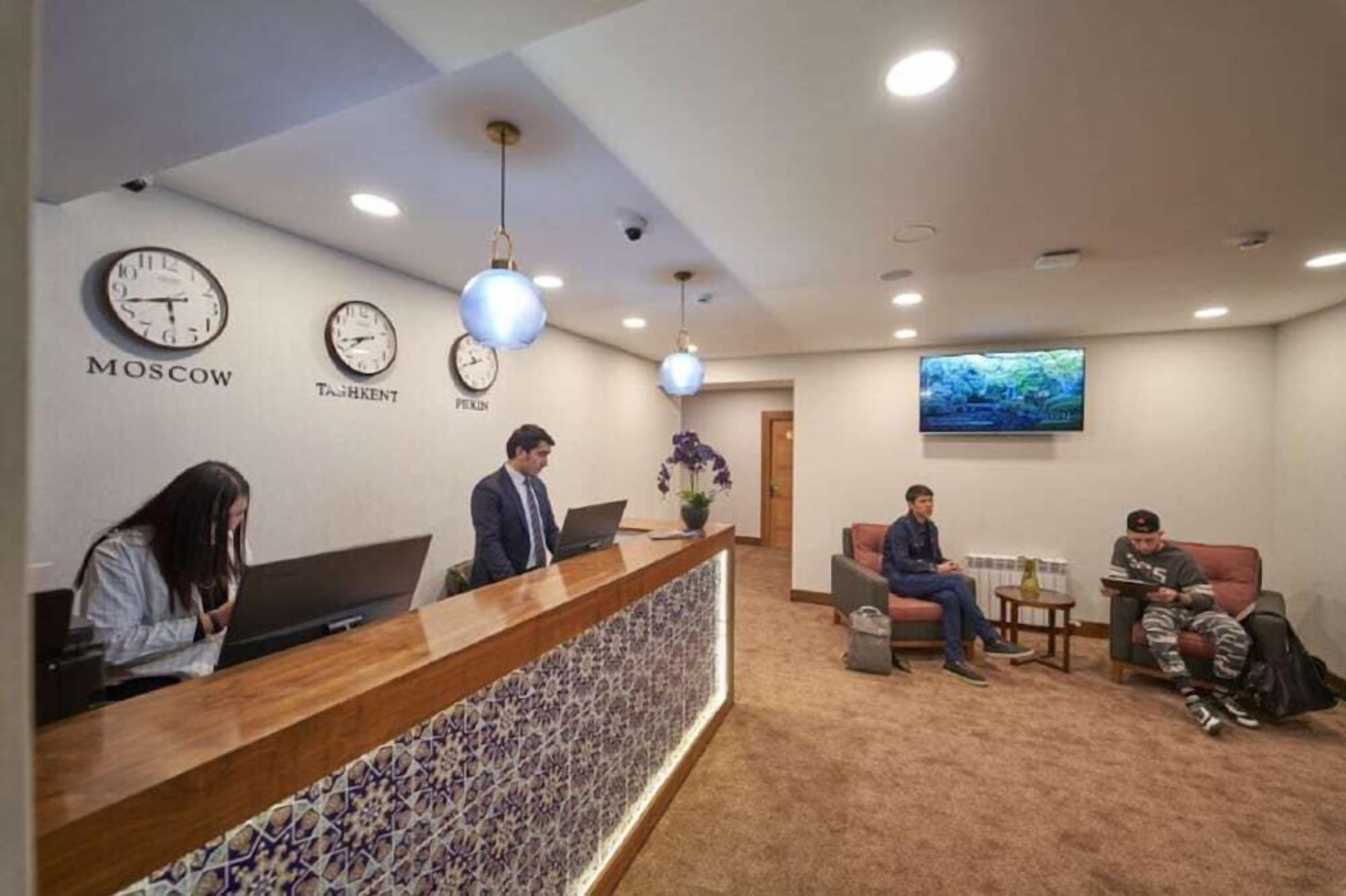 Uma área de recepção com balcão, relógios marcando os horários de Moscovo, Tashkent e Teerão, e uma tela de televisão na parede.