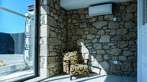 Quarto com paredes de pedra, uma poltrona estampada, uma janela e um aparelho de ar condicionado.