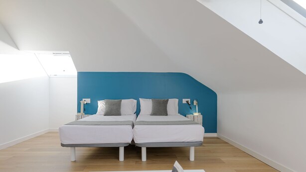 Quarto moderno com teto inclinado, duas camas com roupa de cama branca e almofadas cinzas, uma mesa branca com um controle remoto e uma parede azul decorativa.