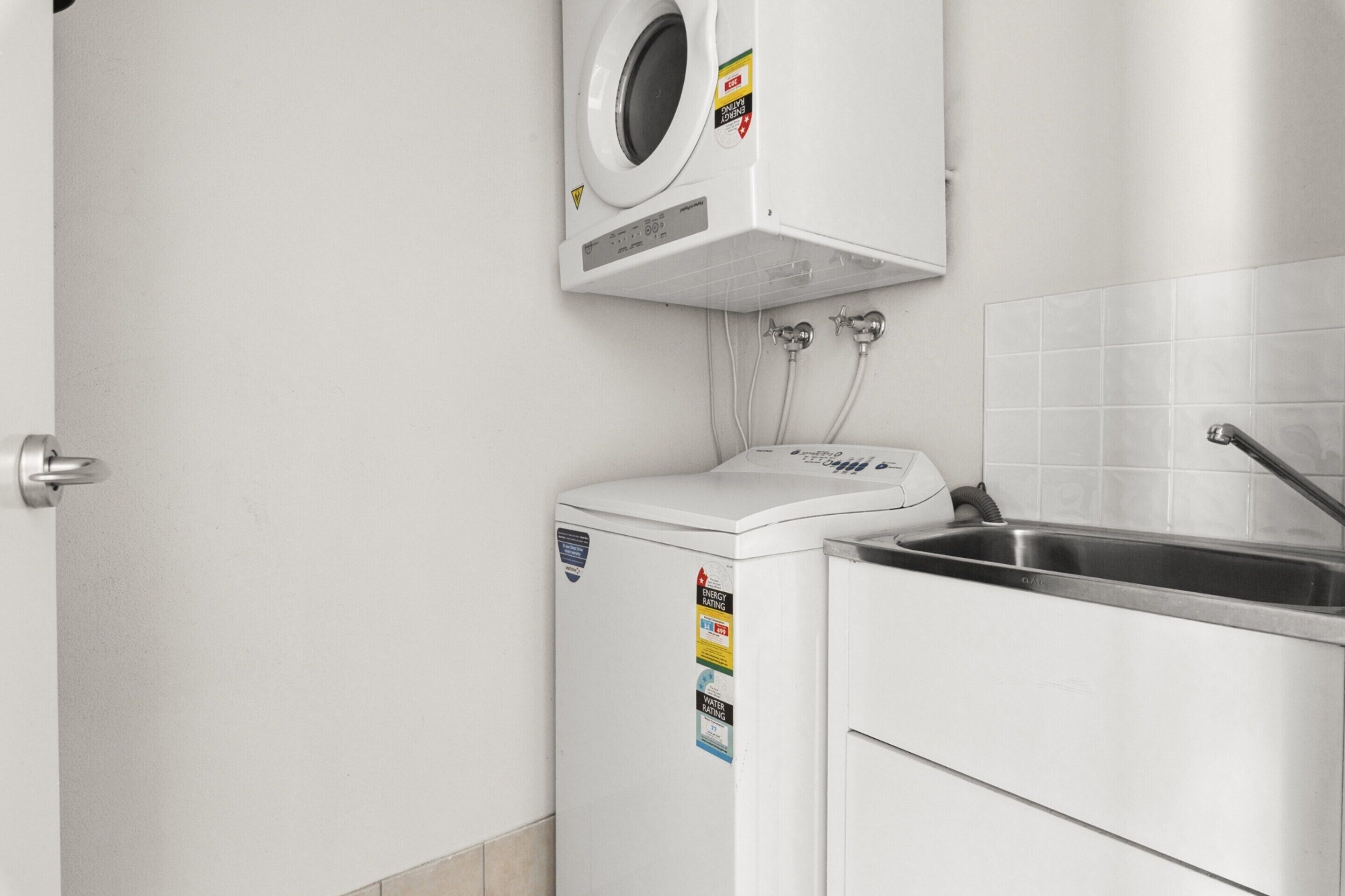 Uma área de lavanderia compacta, com máquina de lavar e secadora, pia de aço inoxidável e painel de azulejos.