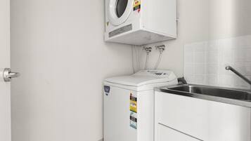 Uma área de lavanderia compacta, com máquina de lavar e secadora, pia de aço inoxidável e painel de azulejos.