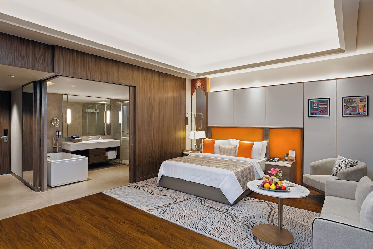 Quarto de hotel moderno com uma cama grande, um espaço de estar, uma mesa pequena com frutas, um banheiro com chuveiro e um espelho grande.
