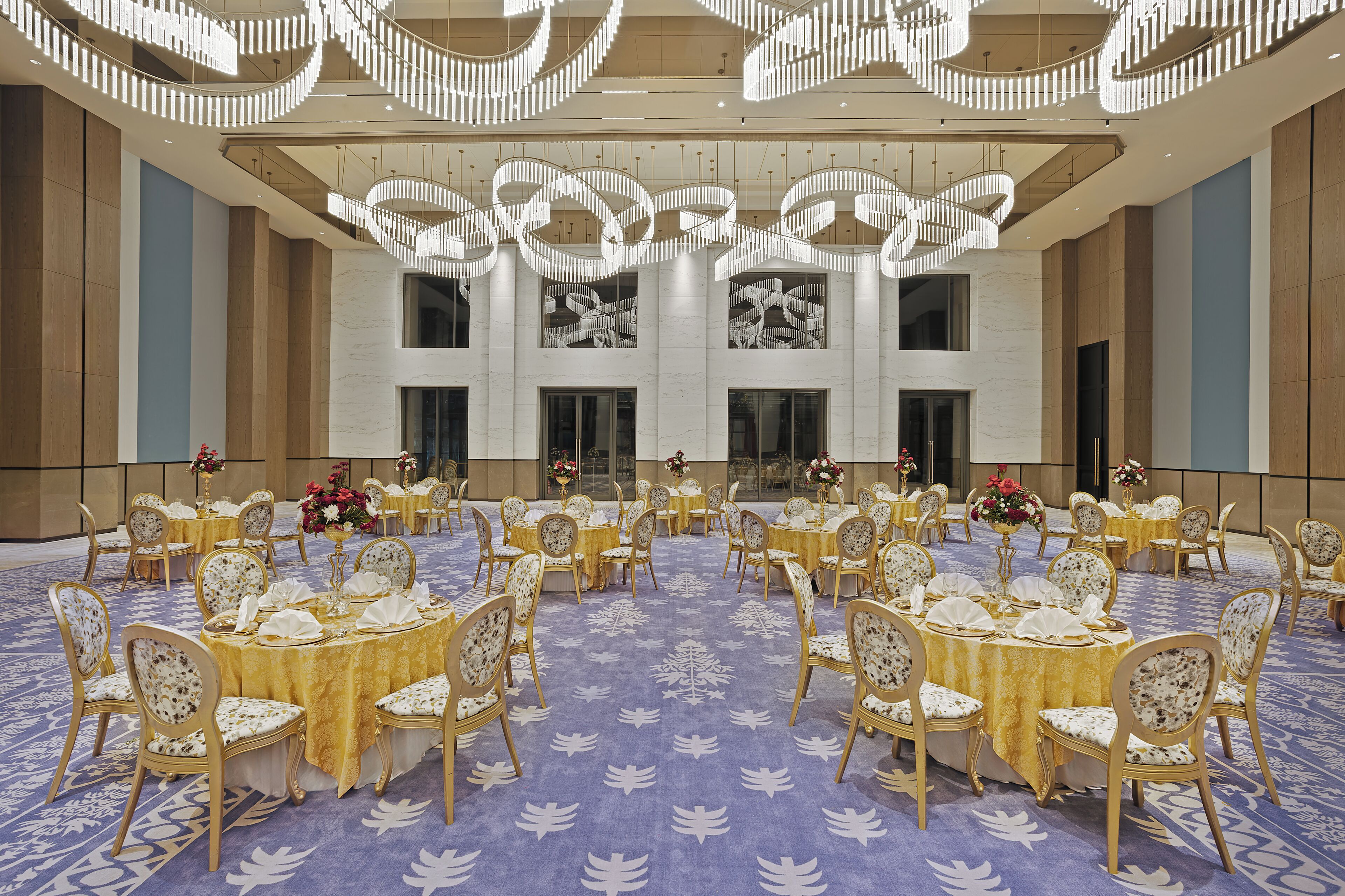 Um amplo salão de banquetes com mesas redondas, cadeiras e um lustre central.