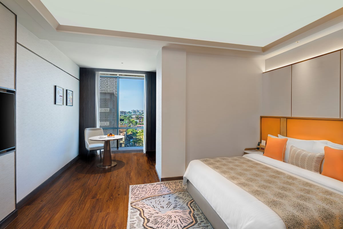 Quarto de hotel moderno com uma cama grande, uma mesinha e vista para a cidade.