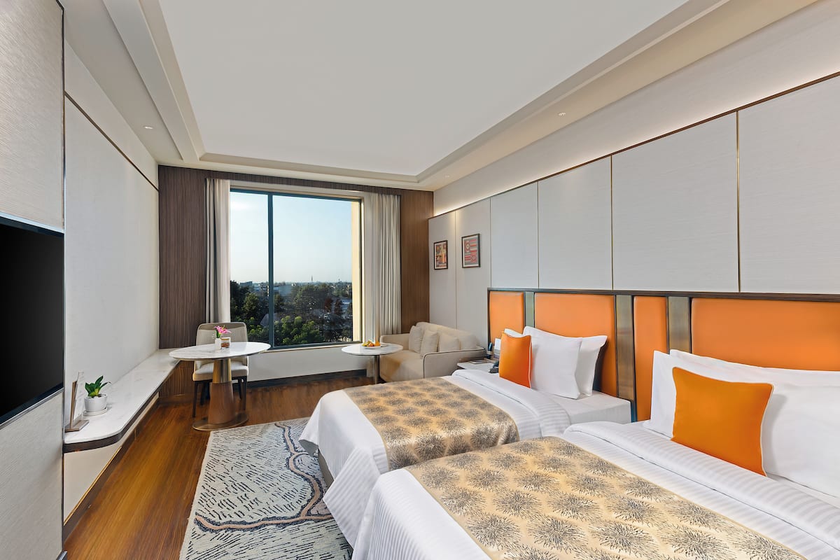 Quarto de hotel com duas camas, uma escrivaninha, uma cadeira, uma televisão e uma janela com vista para a cidade.