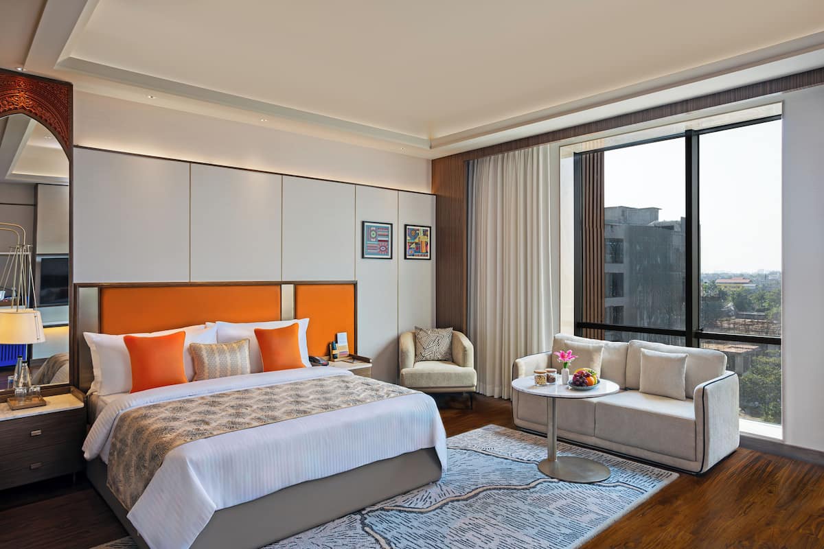 Quarto de hotel com uma cama grande, um sofá, uma cadeira, uma mesinha e uma janela ampla com vista para a cidade.