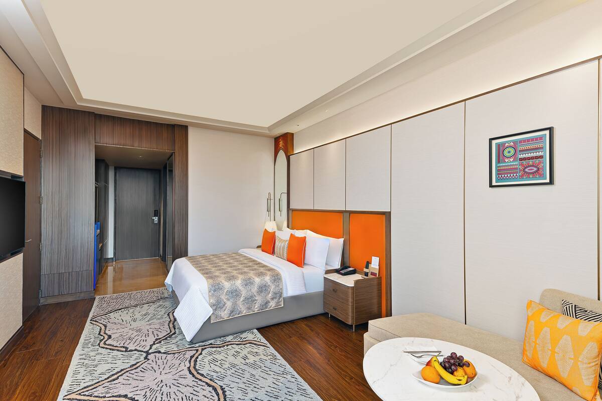 Quarto de hotel moderno com cama, mesa de cabeceira e uma mesa redonda com frutas.