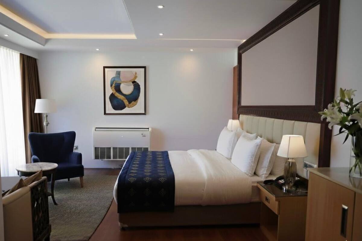 Quarto de hotel com cama, mesas de cabeceira, uma cadeira, escrivaninha e uma grande pintura abstrata na parede.