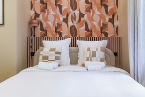 Uma cama bem arrumada com roupa de cama branca e duas almofadas, cabeceira com padrão geométrico e uma luminária fixada na parede.