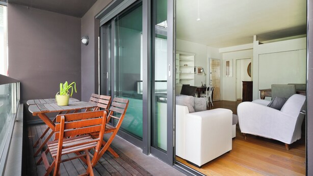 Uma varanda com mesa e cadeiras de madeira, um vaso de planta e uma porta de vidro deslizante que dá para uma sala de estar com sofá branco e uma pequena mesa.
