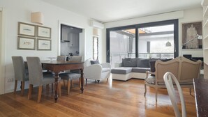 Uma sala de estar espaçosa com piso de madeira, uma mesa de jantar com cadeiras, um sofá e uma grande janela que oferece muita luz natural.