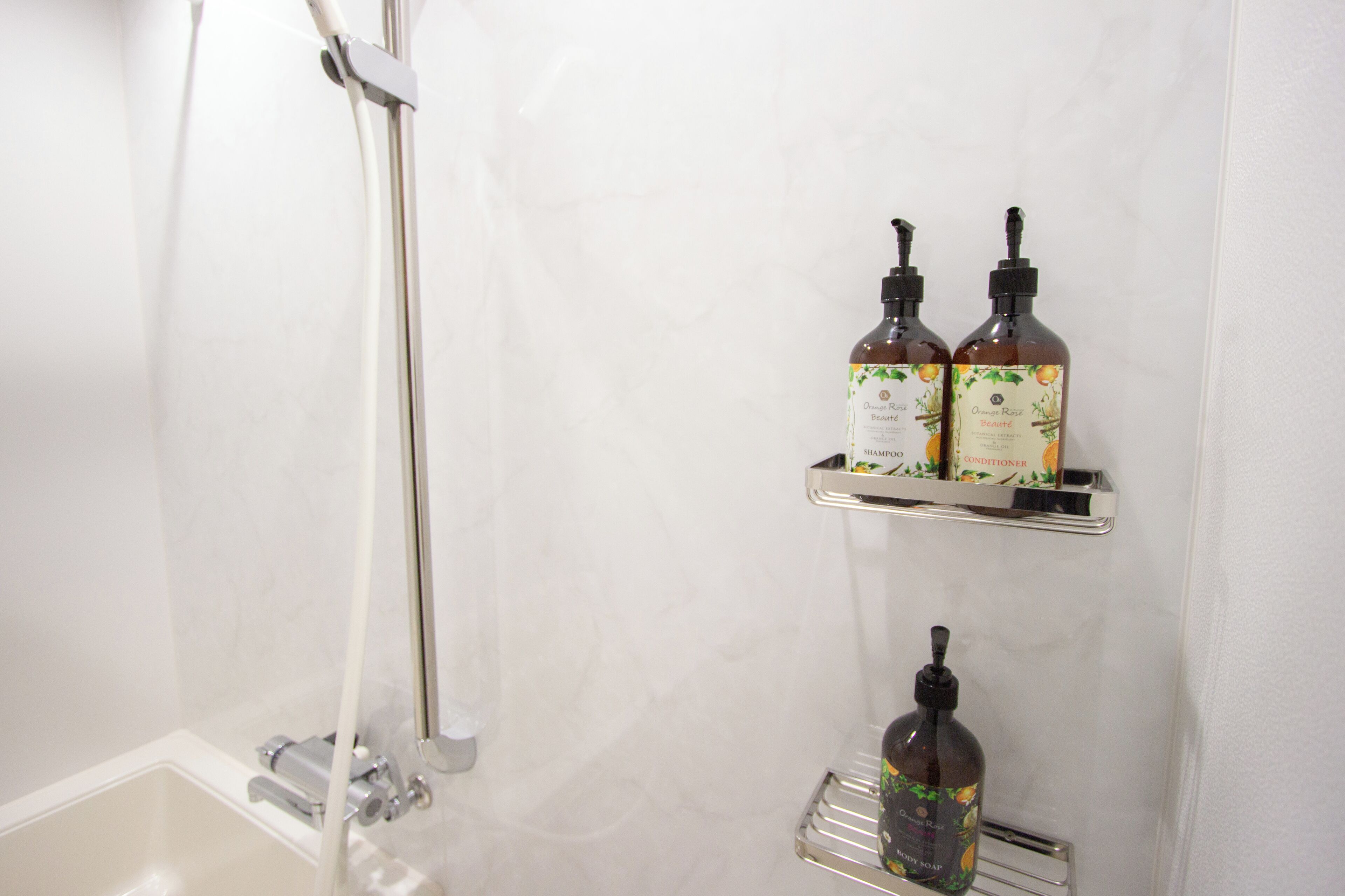 Casa de banho com chuveiro, duas prateleiras com shampoo e condicionador, e uma embalagem de sabonete líquido.