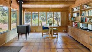 Interior acolhedor de uma cabana com teto de madeira, uma mesa de jantar com cadeiras, uma estante de livros e uma grande janela com vista para o exterior.