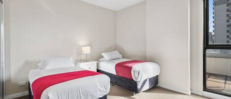 Quarto de hotel com duas camas, uma mesinha de cabeceira, uma luminária e uma janela grande com vista para a cidade.