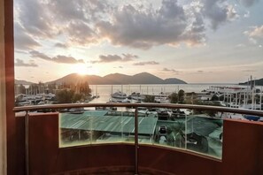 Uma varanda com vista para um porto de iates e montanhas ao pôr do sol.