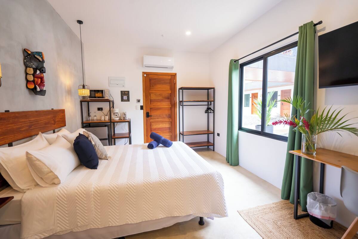 Quarto de hotel com cama, porta de madeira, prateleira com planta e janela com cortinas.