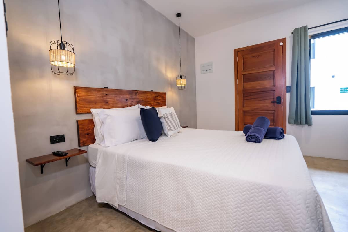 Quarto com cabeceira de madeira, colcha branca acolchoada, duas luminárias de cabeceira, mesa de cabeceira de madeira e uma janela com cortinas.