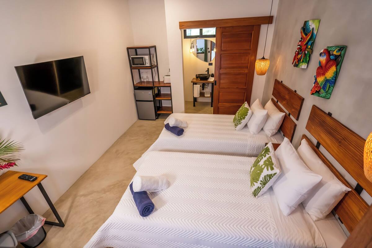 Quarto de hotel com duas camas, televisão, secretária e banheiro visível através de uma porta aberta.