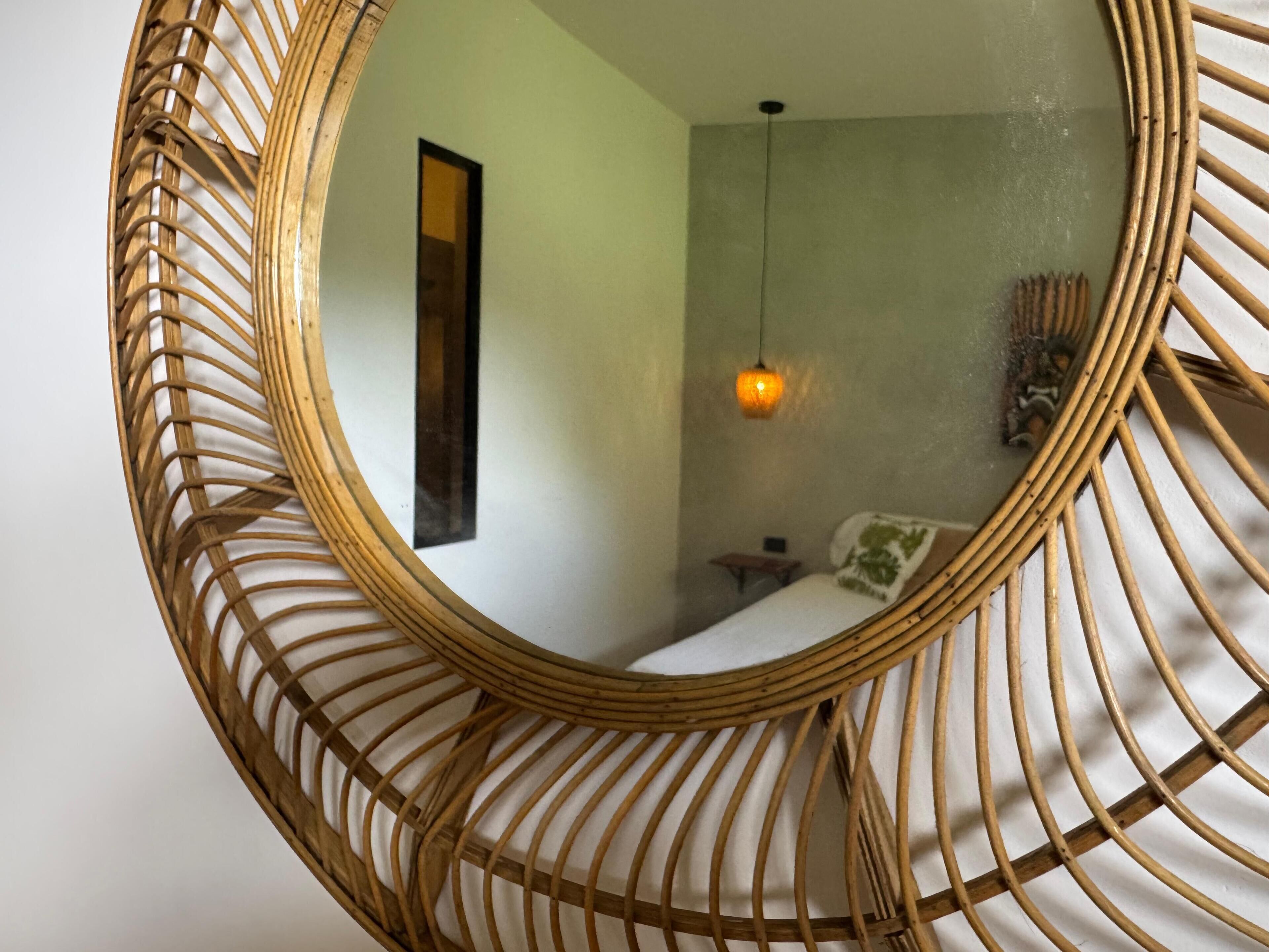 Uma cadeira circular trançada, um espelho redondo, uma cama com colchão branco e almofada verde, um lustre pendente e uma pequena mesa com uma planta.