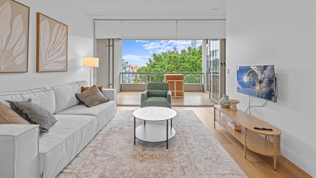 Uma sala de estar moderna com um sofá branco, uma mesa de centro redonda e uma poltrona verde. Uma televisão de tela plana está montada na parede, e há uma varanda com vista para árvores e edifícios.