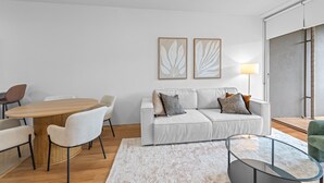 Uma sala de estar moderna com uma mesa redonda de madeira, sofá branco e mesa de centro de vidro. Há duas obras de arte com folhas emolduradas na parede e um abajur.