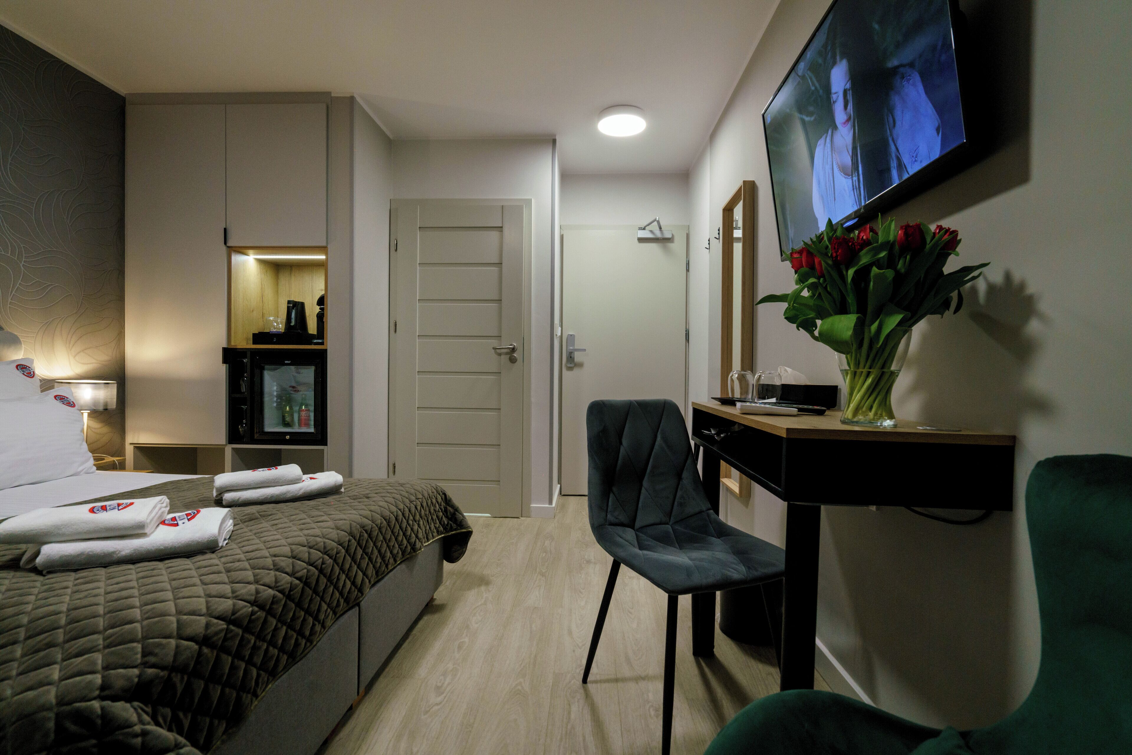 Quarto de hotel com cama, escrivaninha, cadeira, televisão e um vaso com flores.