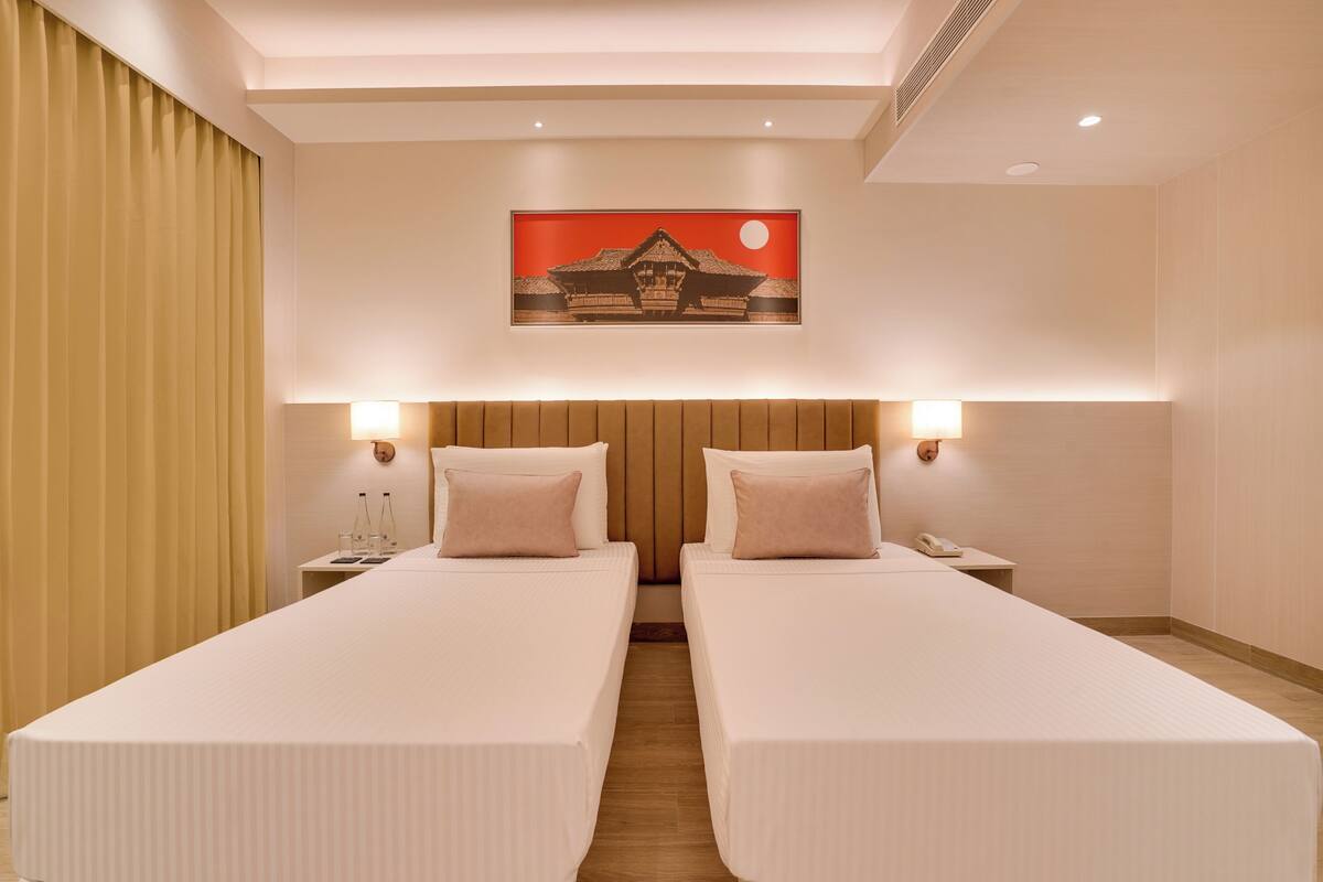 Quarto de hotel com duas camas, cabeceira de madeira, luminárias laterais, mesa de cabeceira com telefone e pintura de um edifício tradicional.