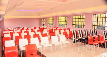 Uma sala de conferências com fileiras de cadeiras, janelas amplas e um logotipo na parede.