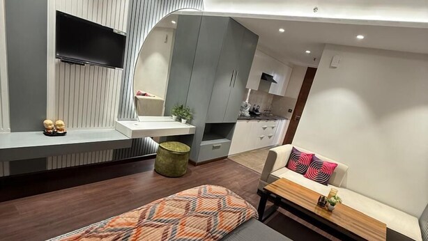 Quarto de hotel moderno com cama, sofá, pequena mesa, TV fixada na parede e área de cozinha ao fundo.