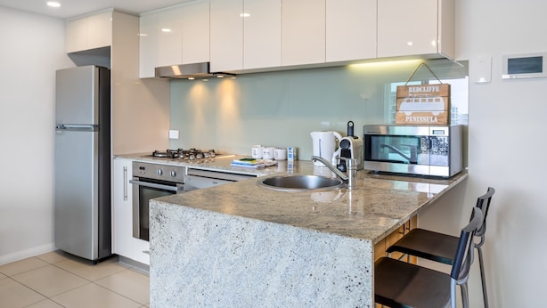 Uma cozinha moderna com armários brancos, eletrodomésticos de aço inoxidável, bancada de granito e uma placa com a inscrição "REDCLIFFE PENINSULA".