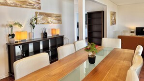 Uma sala de jantar moderna com uma mesa de vidro, cadeiras estofadas brancas e um aparador de madeira com uma lâmpada e objetos decorativos.