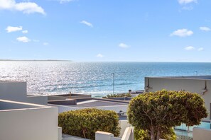 Vista costeira com céu claro, mar calmo e edifícios modernos com telhados planos e vegetação.