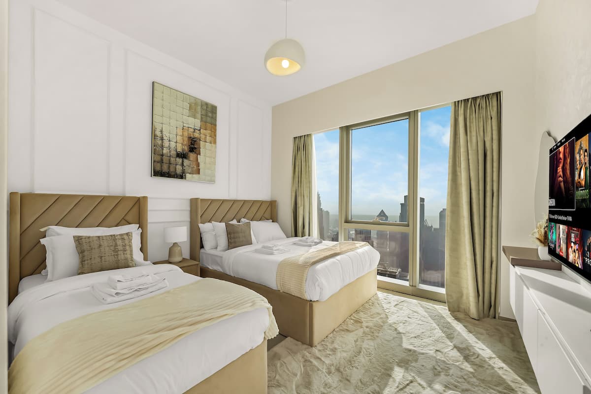 Quarto de hotel com duas camas, uma janela grande com vista para a cidade, uma televisão de tela plana e um quadro na parede.