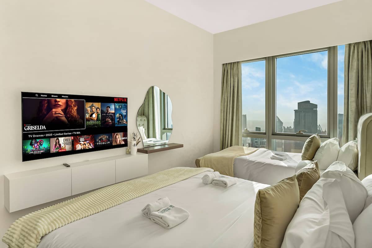 Quarto de hotel com uma televisão grande exibindo a interface Netflix, uma cama com roupa de cama branca, uma mesinha de cabeceira com abajur e vista para a cidade através de grandes janelas.