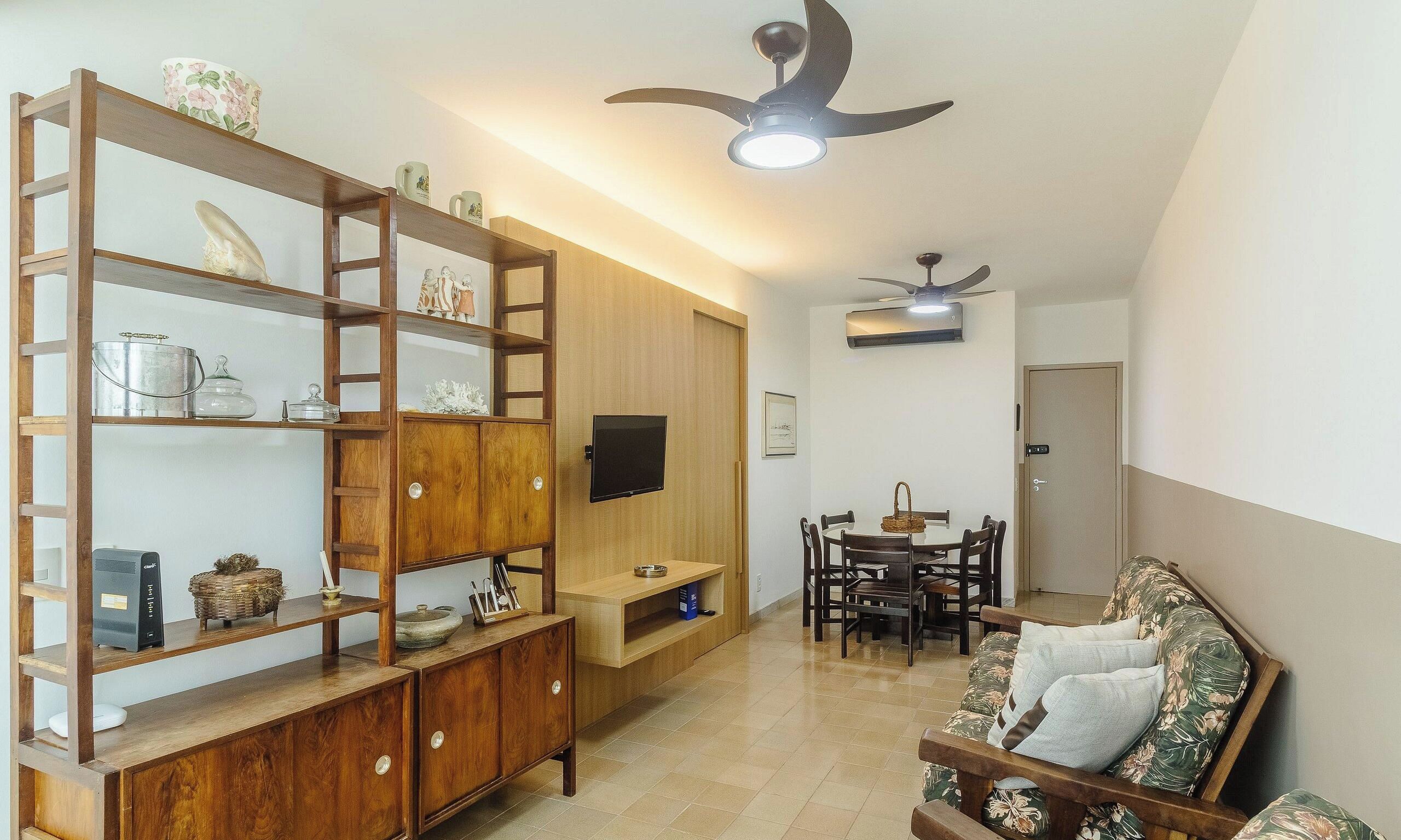 Uma sala de estar com um armário de madeira, uma televisão, um ventilador de teto e um espaço de jantar com cadeiras e uma mesa.