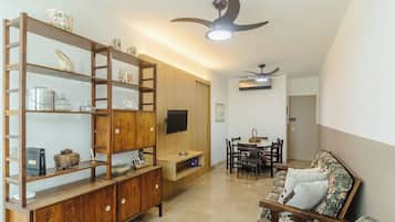 Uma sala de estar com um armário de madeira, uma televisão, um ventilador de teto e um espaço de jantar com cadeiras e uma mesa.