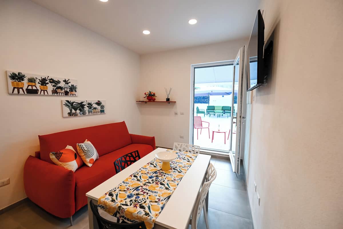 Uma sala de estar moderna com um sofá vermelho, uma mesa de jantar com toalha colorida e uma grande janela com vista para móveis de exterior.