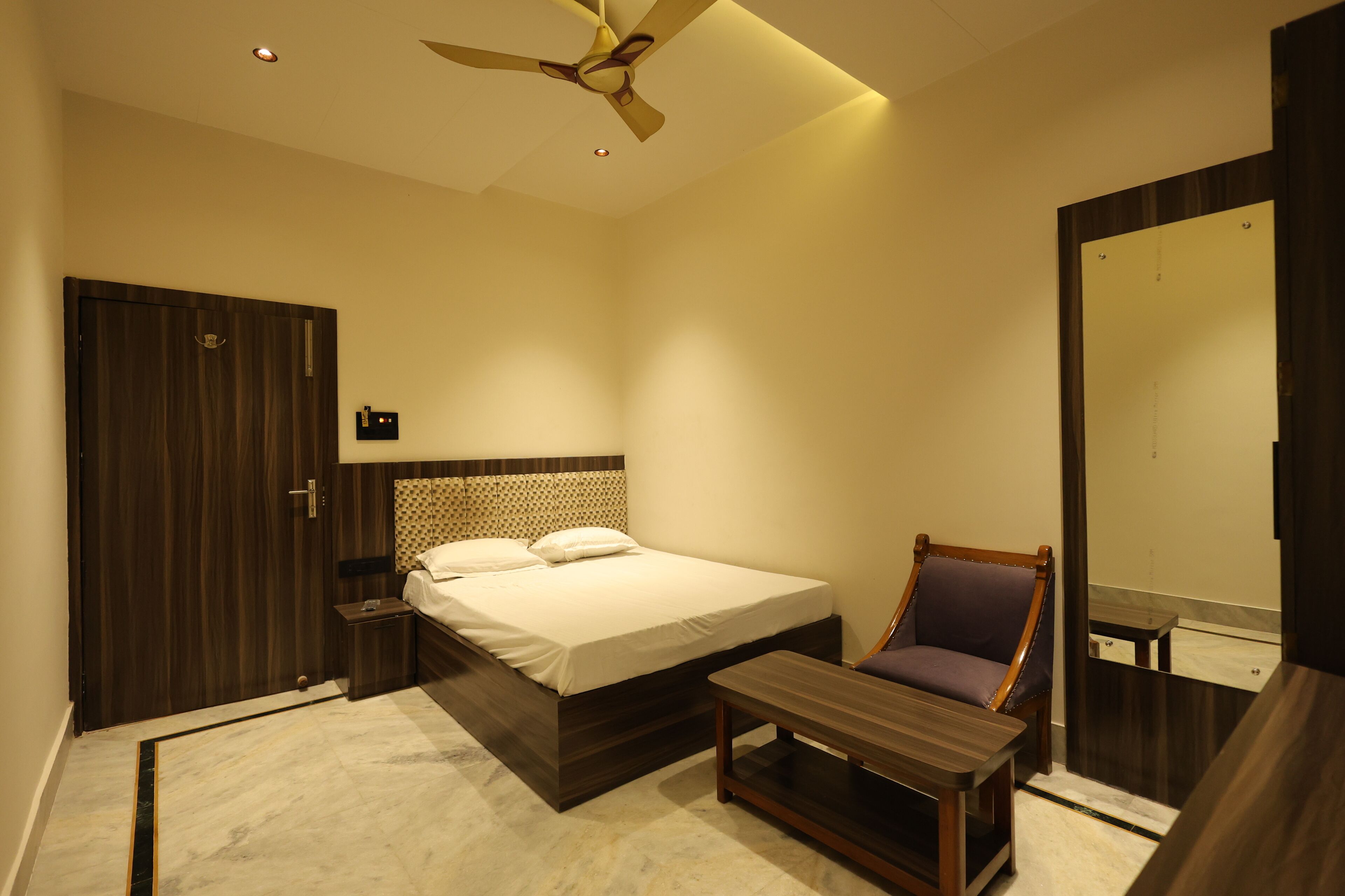 Quarto de hotel com uma cama, uma cadeira, uma mesa pequena, um ventilador de teto e um espelho.