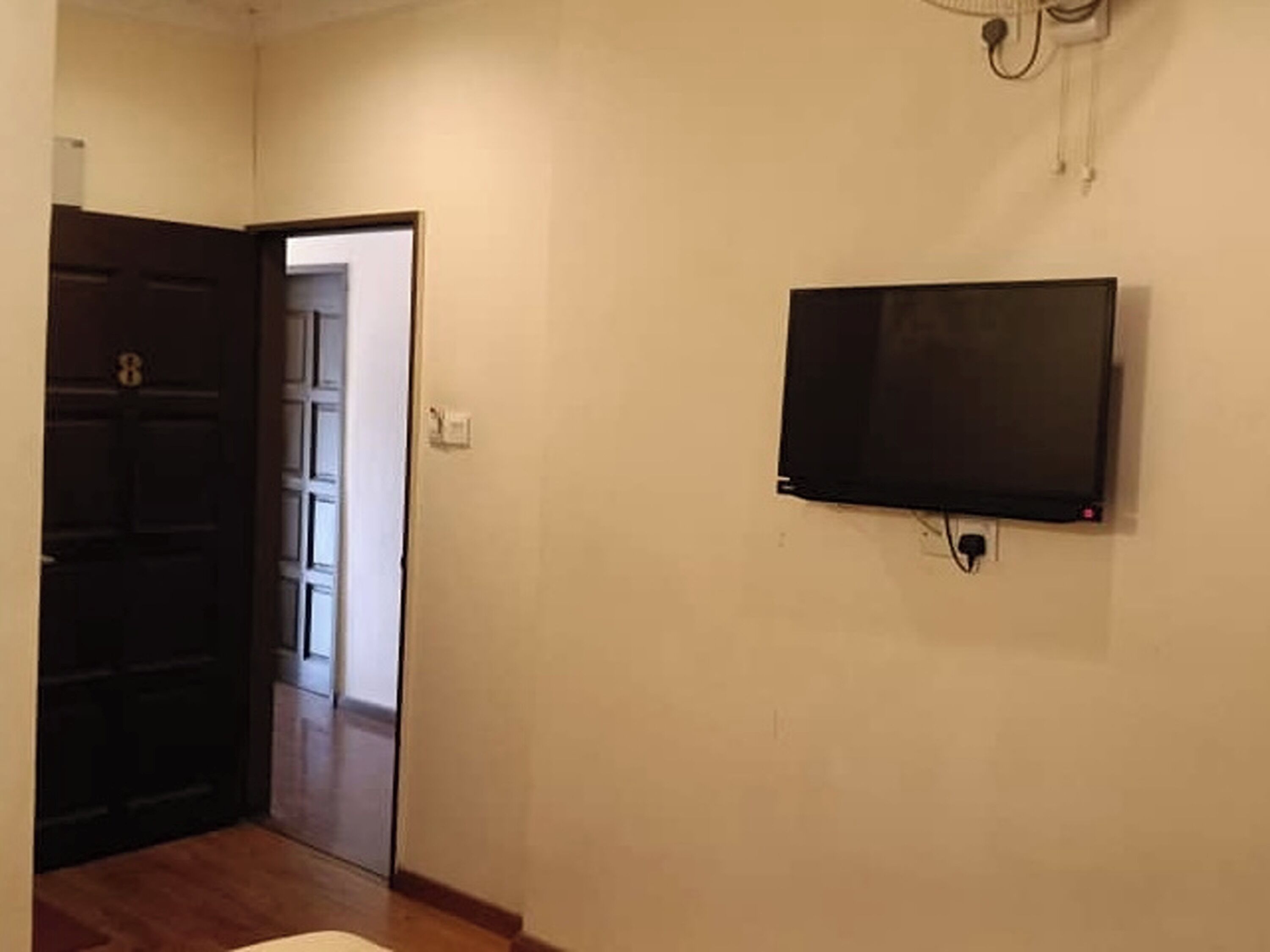 Um quarto com televisão fixada na parede, uma porta que dá para outro espaço e piso de madeira.