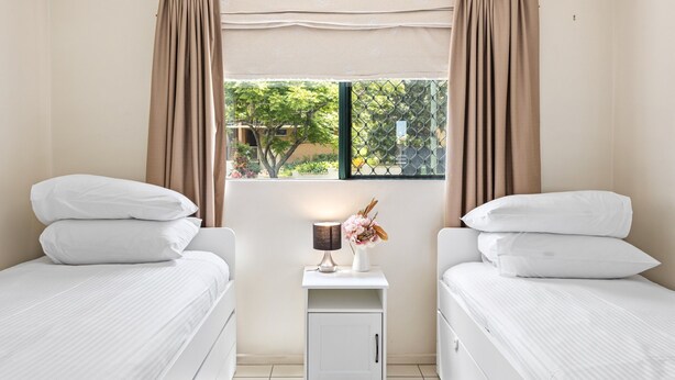 Quarto com duas camas de solteiro, mesa de cabeceira com abajur e vaso, e janela com cortinas.